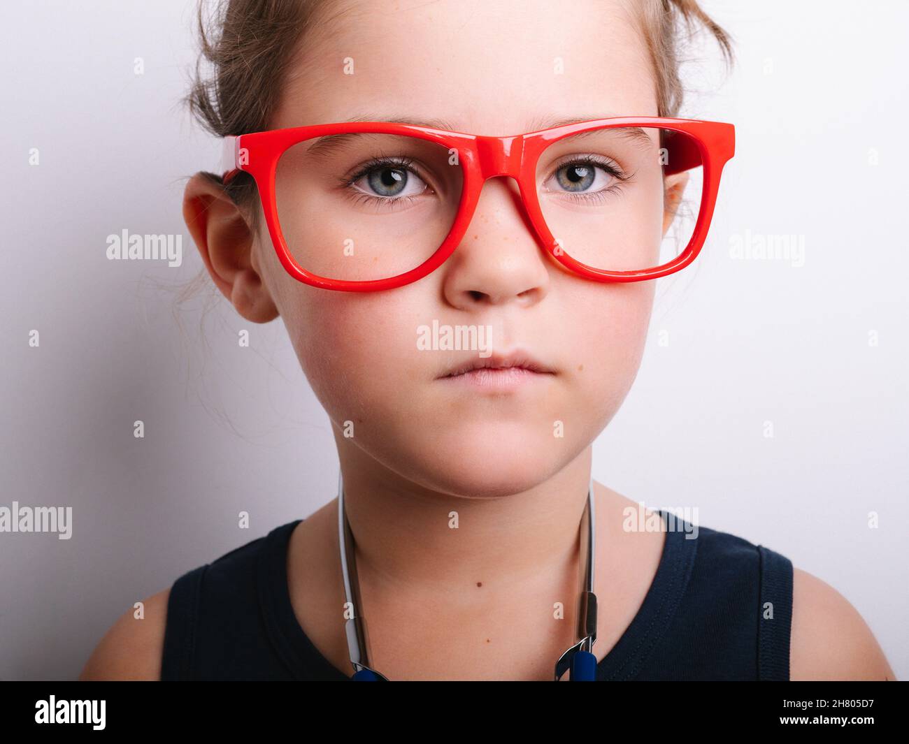 girl in red eyeglasses with stethoscope while looking at camera and playing doctor on white ...