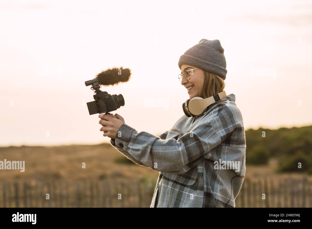 Side view of young female blogger in hat smiling shooting video on ...
