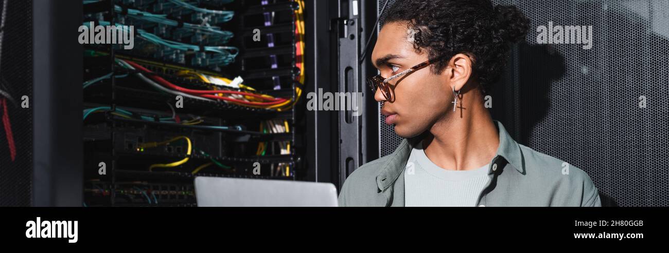 african american engineer with laptop looking at wires in server while working in data center ...