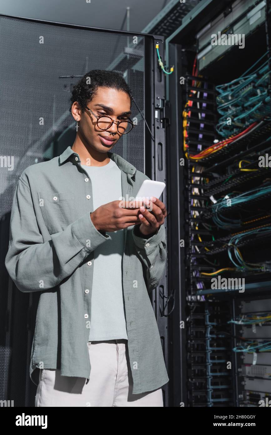 smiling african american programmer using mobile phone while making diagnostics of server in ...