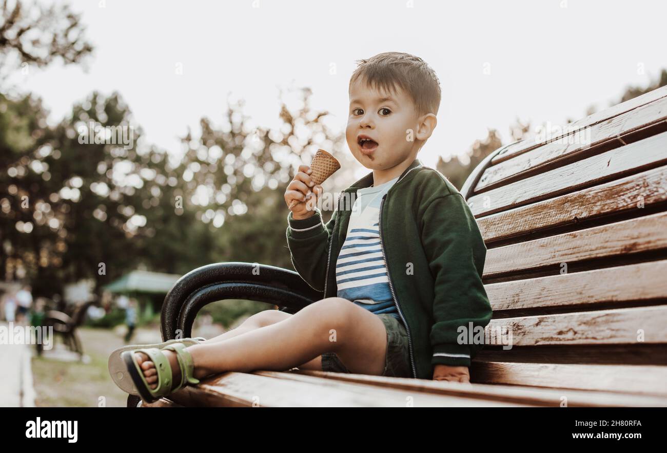 Cucaian little boy eating ice cream sitting on a park bench, looking at ...