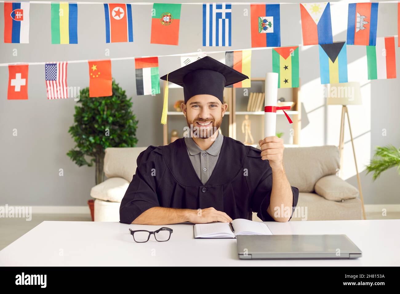 Happy international online university graduate sitting at desk, holding ...