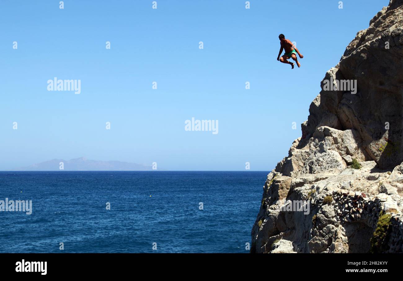 A man jumping in water from a rock at Kamari Beach in Santorini, Greece ...