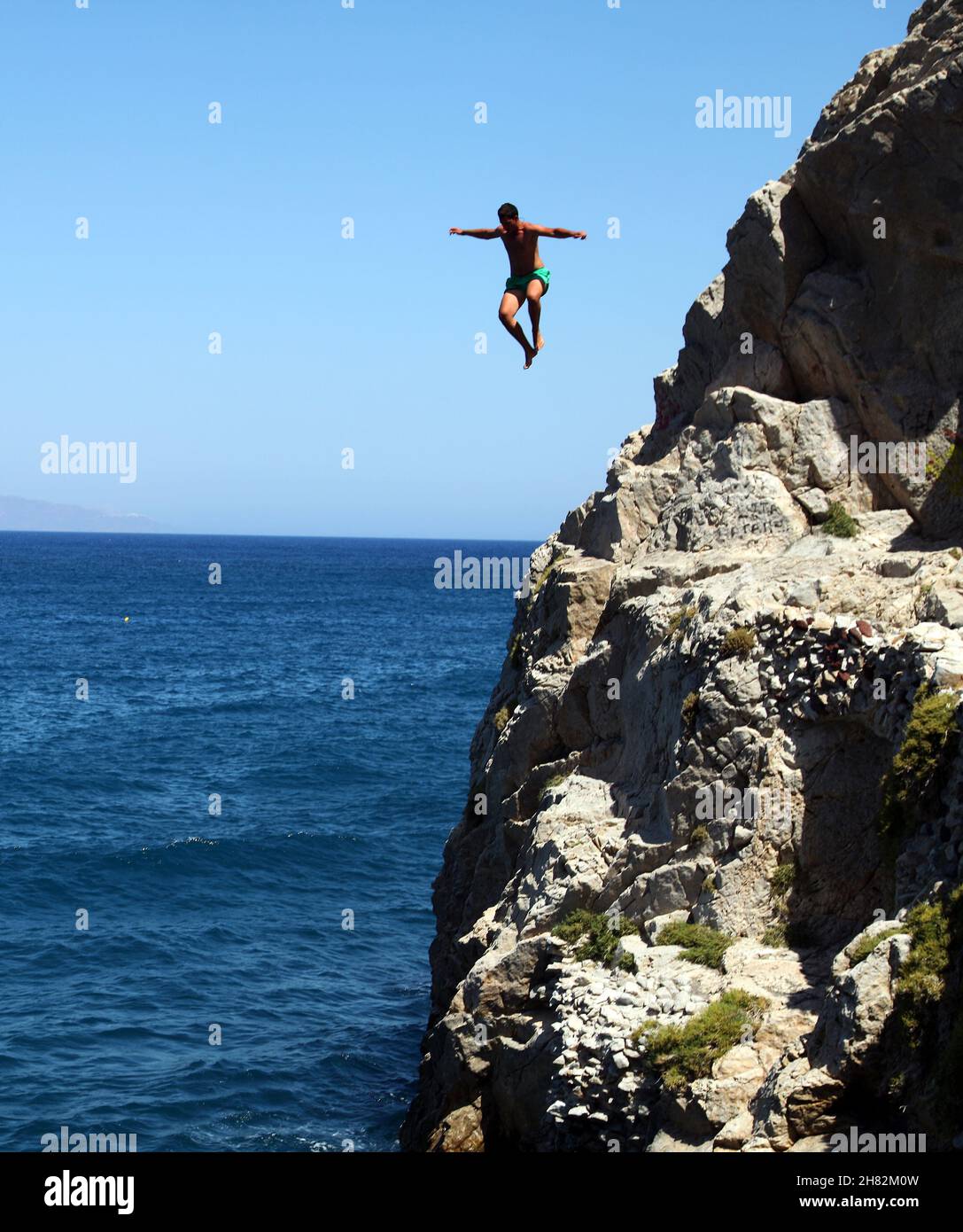 A man jumping in water from a rock at Kamari Beach in Santorini, Greece ...