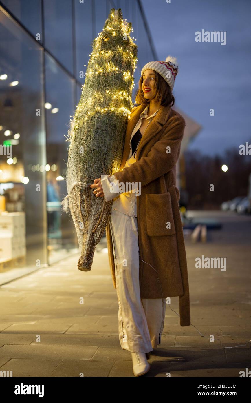 Woman carrying Christmas tree with illuminated garland near the ...