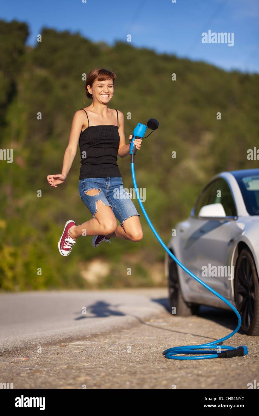 Beautiful young girl jumping next to an electric car. Holding blue ...