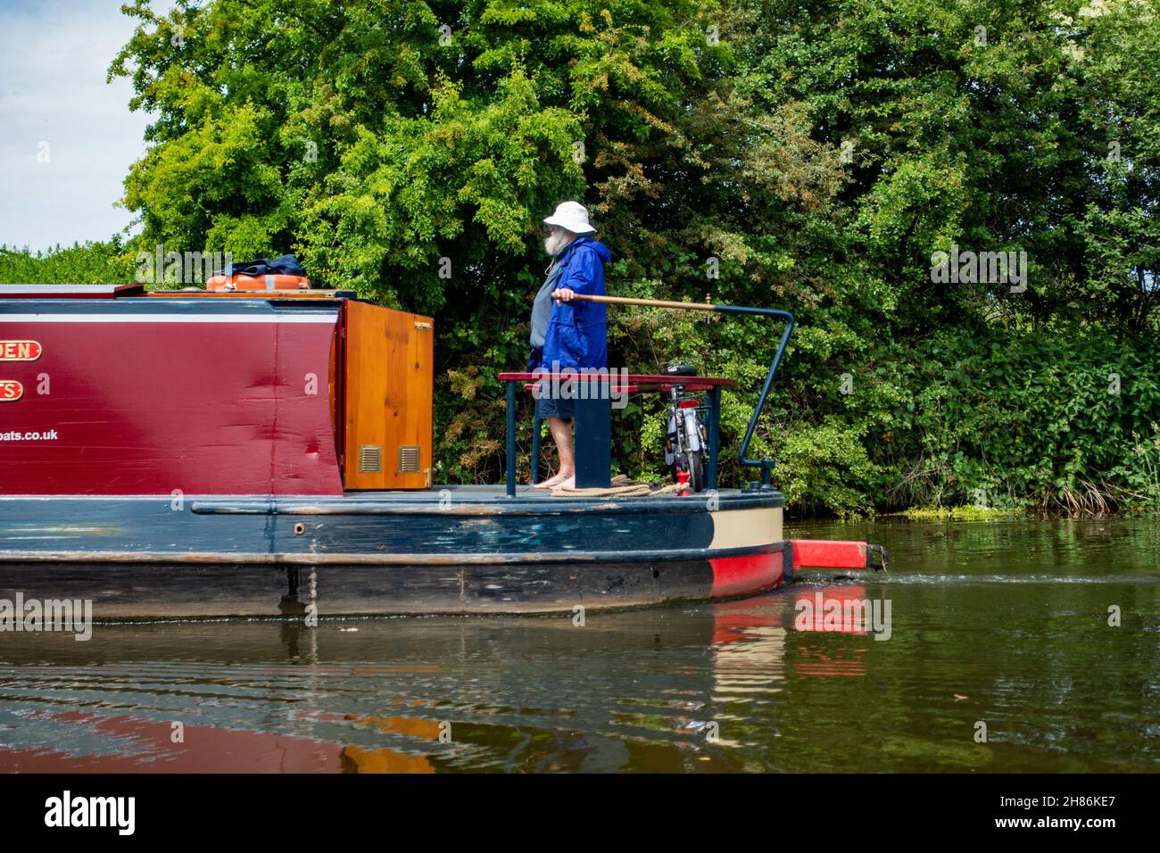 Man standing on the stern of a Red narrowboat using the tiller, sailing ...