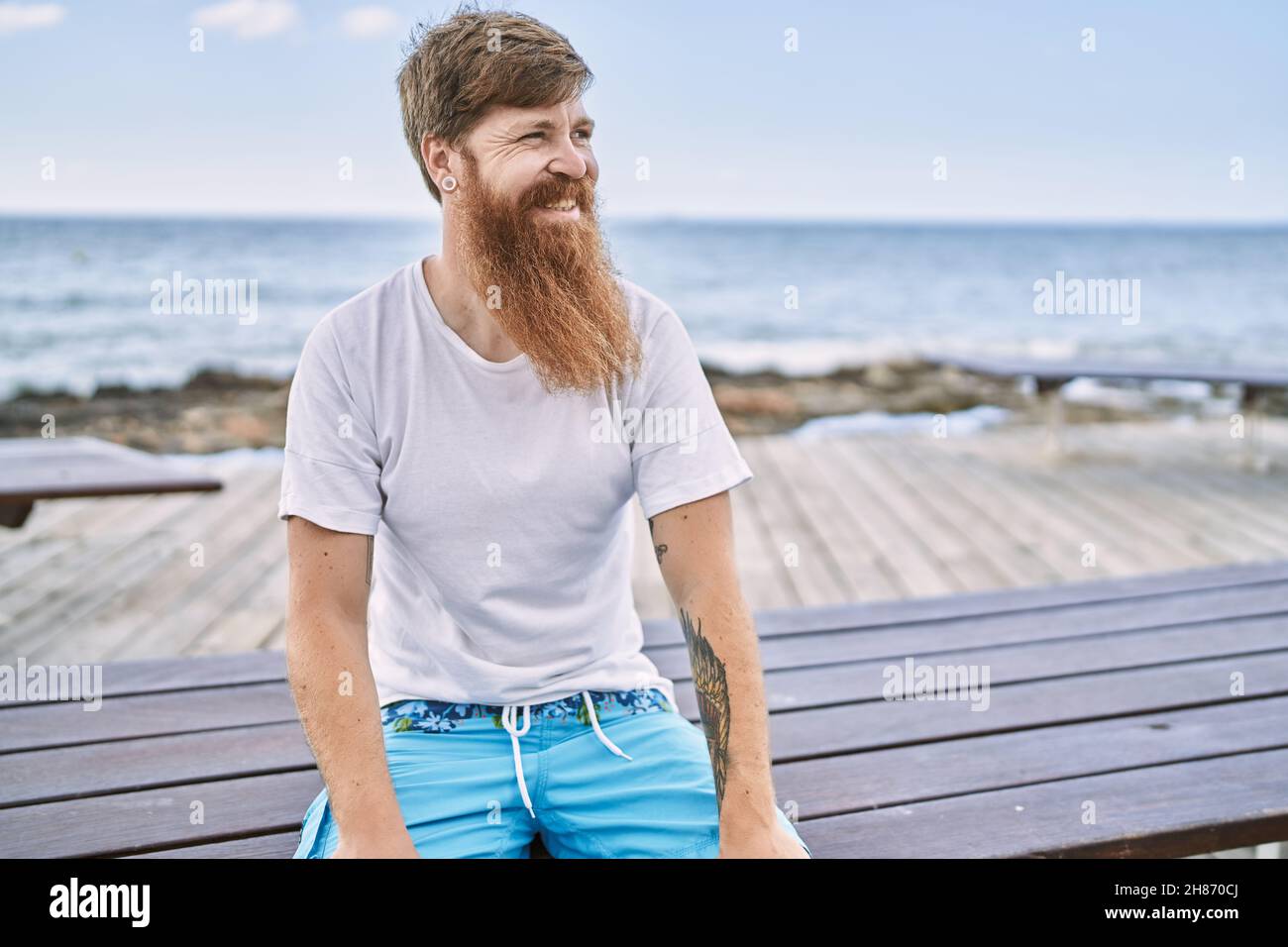 Young redhead man smiling happy sitting on the bench at the beach Stock ...