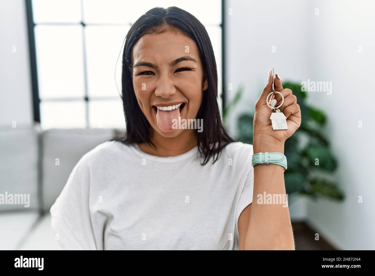 Young hispanic woman holding keys of new home sticking tongue out happy with funny expression ...