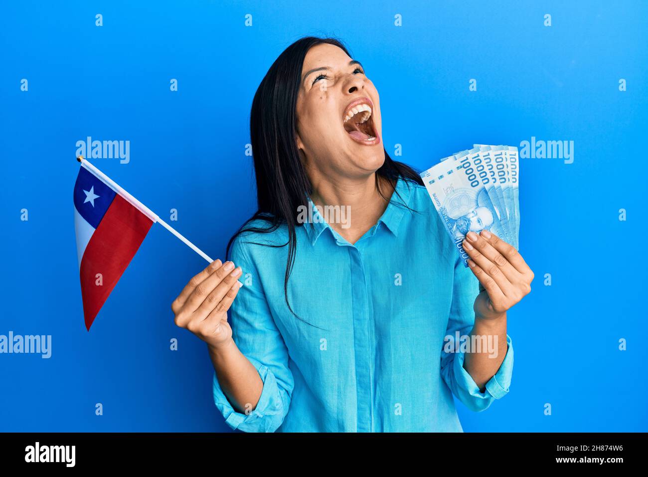 Young latin woman holding chile flag and chilean pesos banknotes angry ...