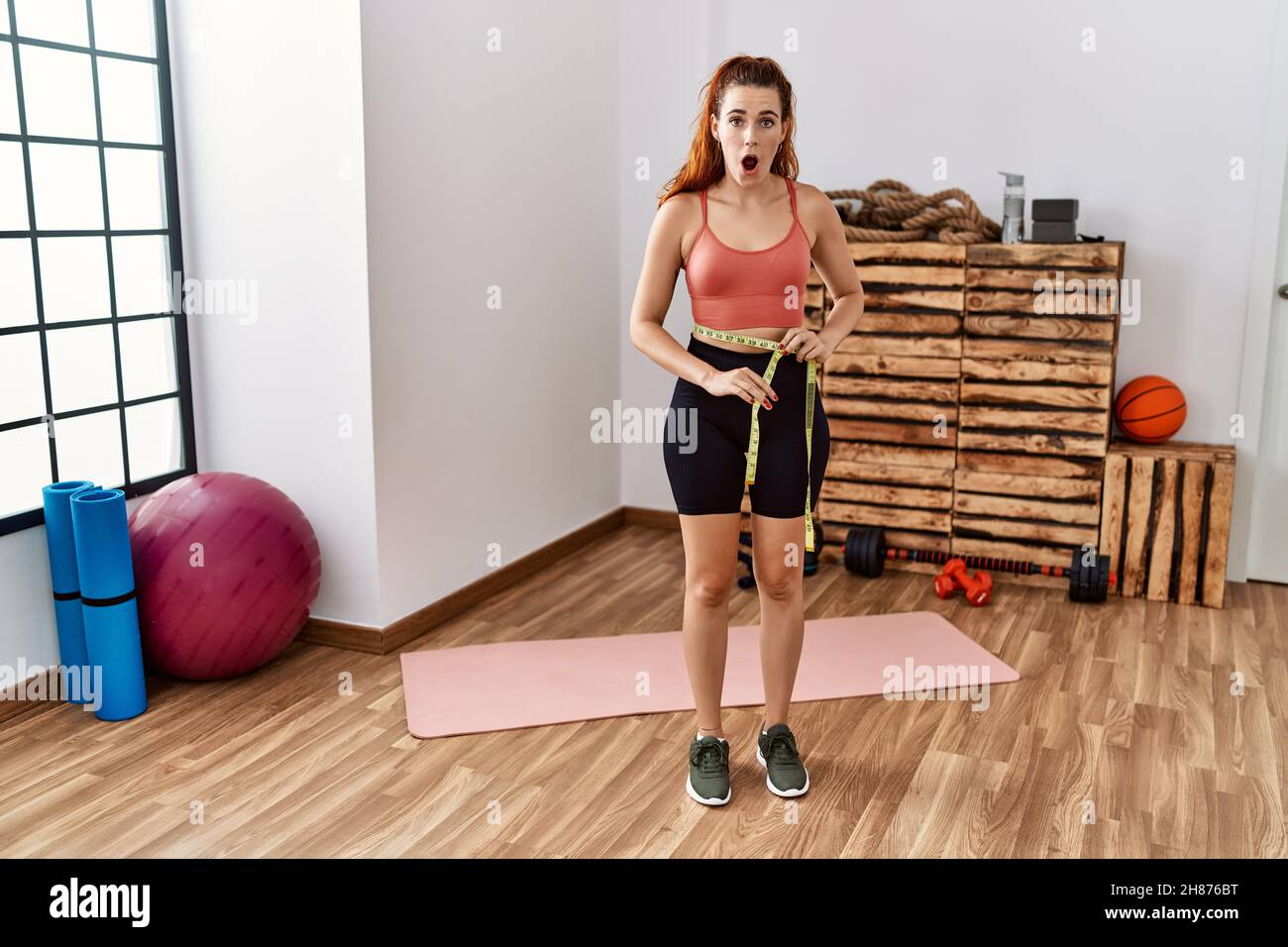 Young redhead woman using tape measure measuring waist at the gym ...