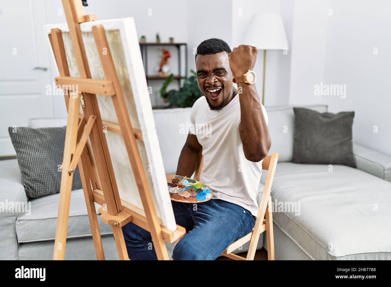 Young african man painting on canvas at home angry and mad raising fist ...