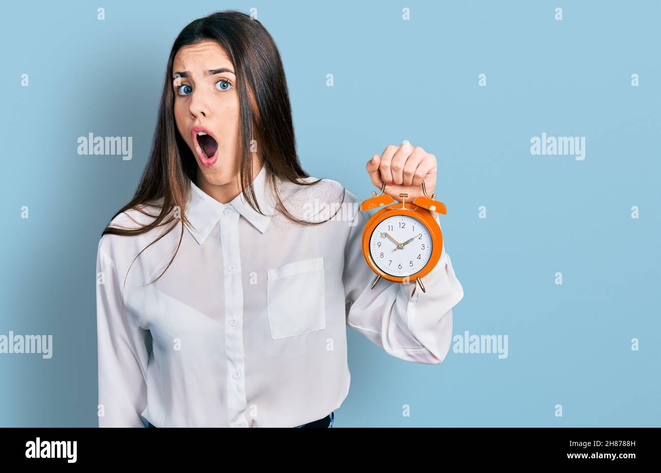 Young brunette teenager holding alarm clock scared and amazed with open ...