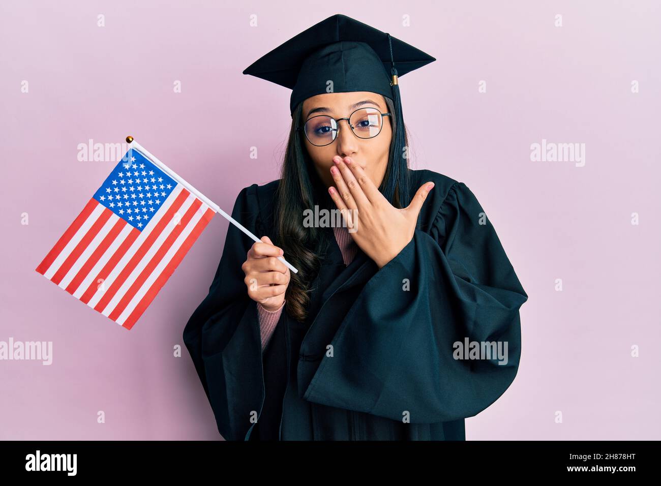 Young hispanic woman wearing graduation cap and ceremony robe holding ...