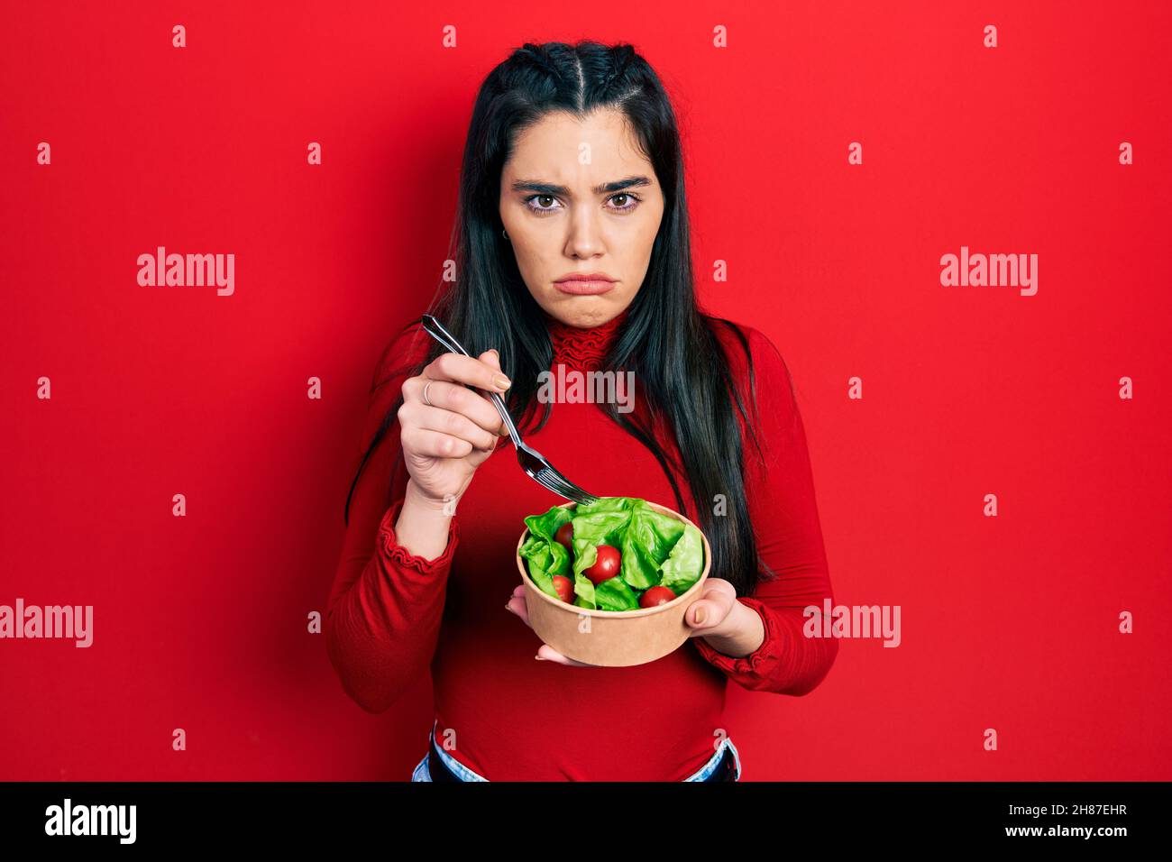 Young hispanic girl eating salad skeptic and nervous, frowning upset ...