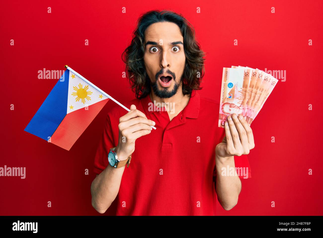 Young hispanic man holding philippines flag and pesos banknotes afraid ...