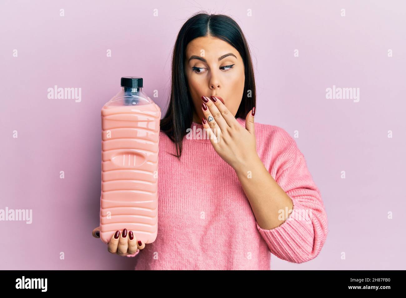 Beautiful brunette woman holding laundry softener bottle covering mouth with hand, shocked and ...