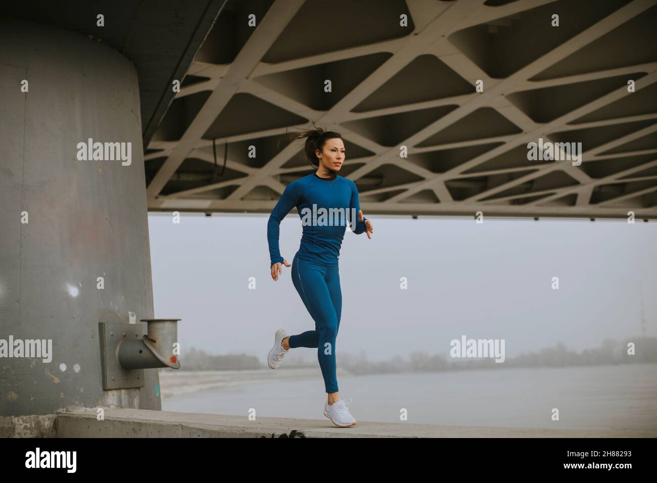 Pretty young woman in blue track suit running by the river at autumn ...