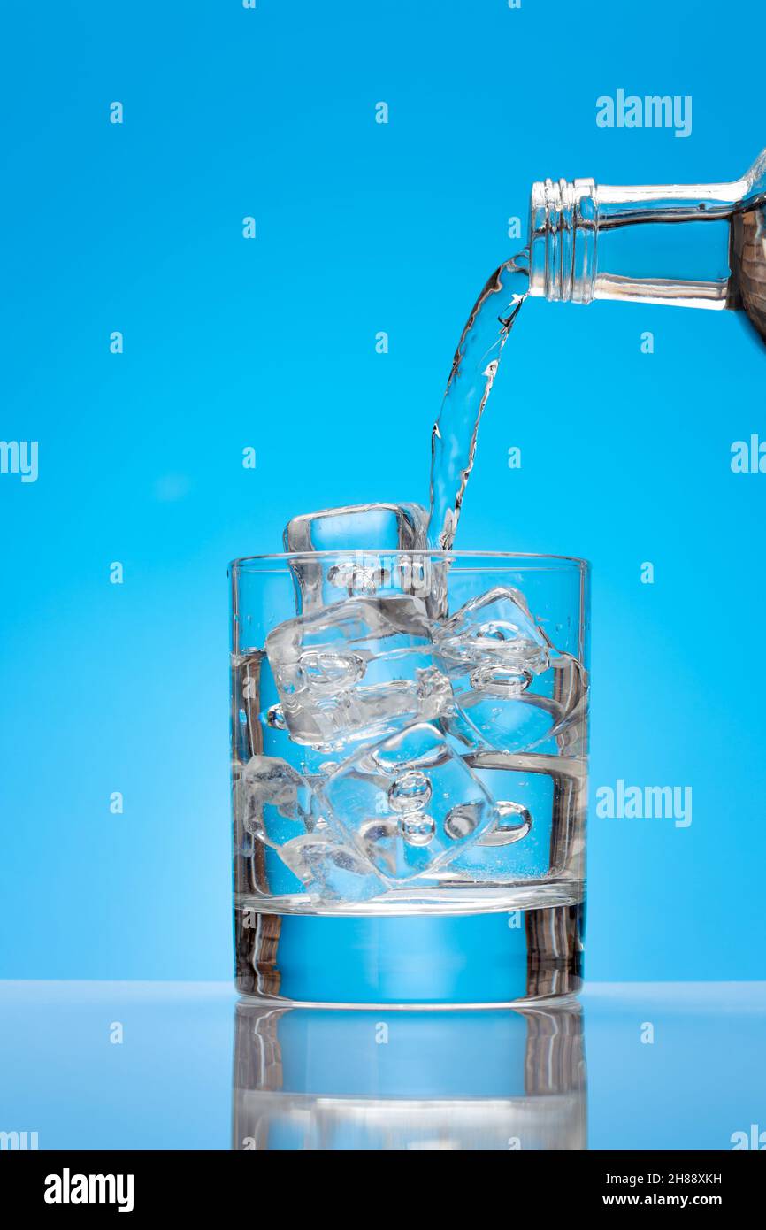 Fresh water pouring from bottle into glass with ice cubes over blue background Stock Photo - Alamy