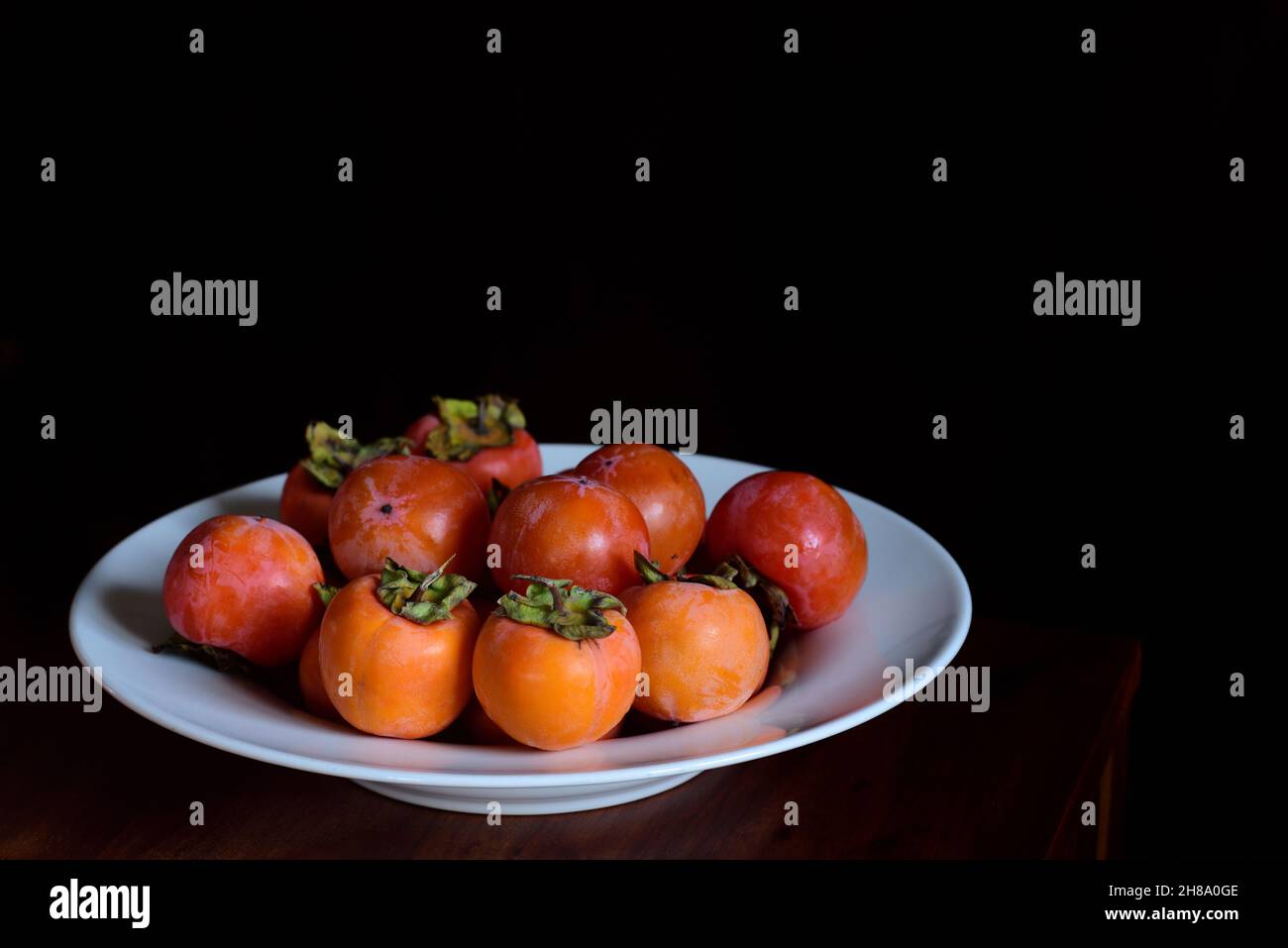A plate of ripe persimmons stands on a wooden table against a dark ...
