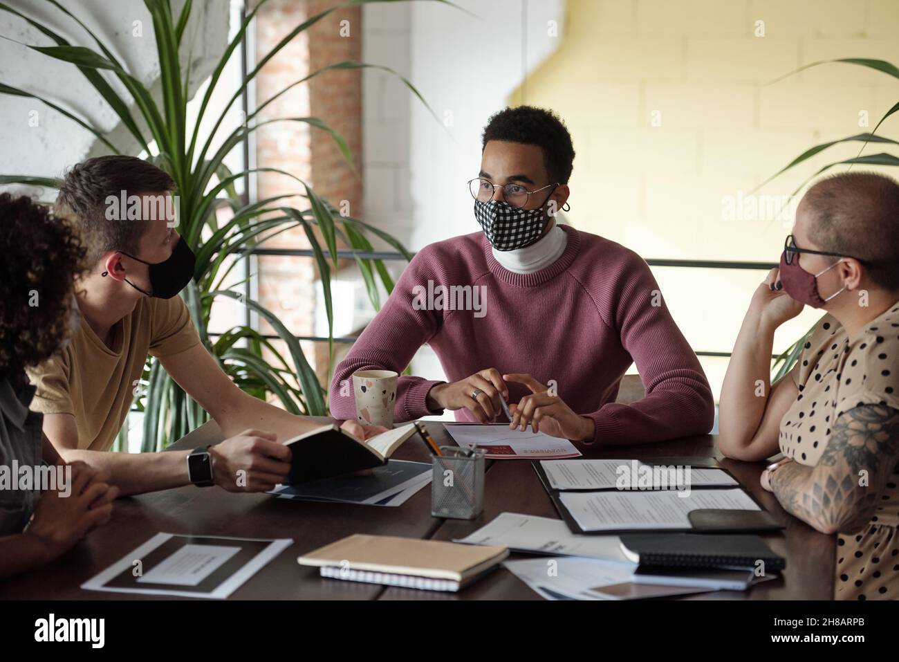 Young confident businessman in mask sitting by table in front of co ...