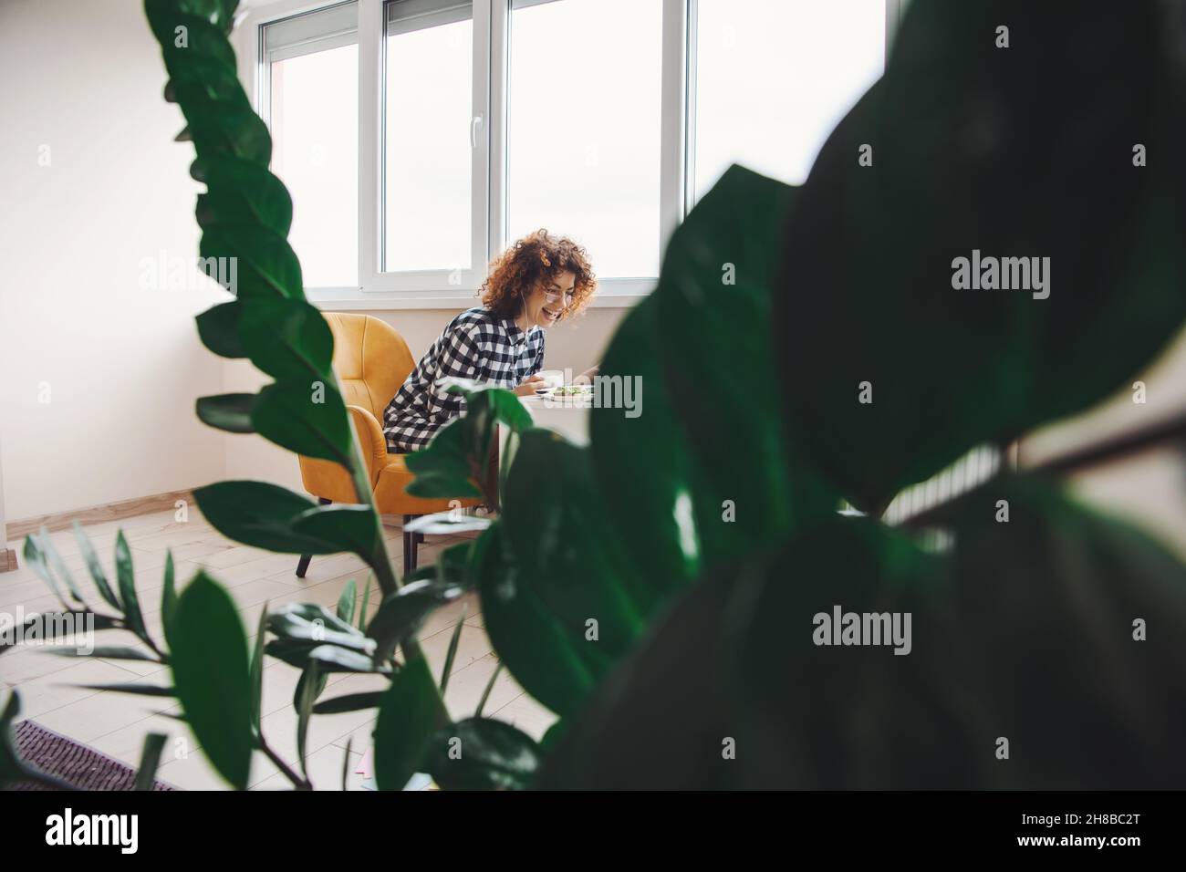 View from behind the leaves of a plant. Woman eating breakfast while ...