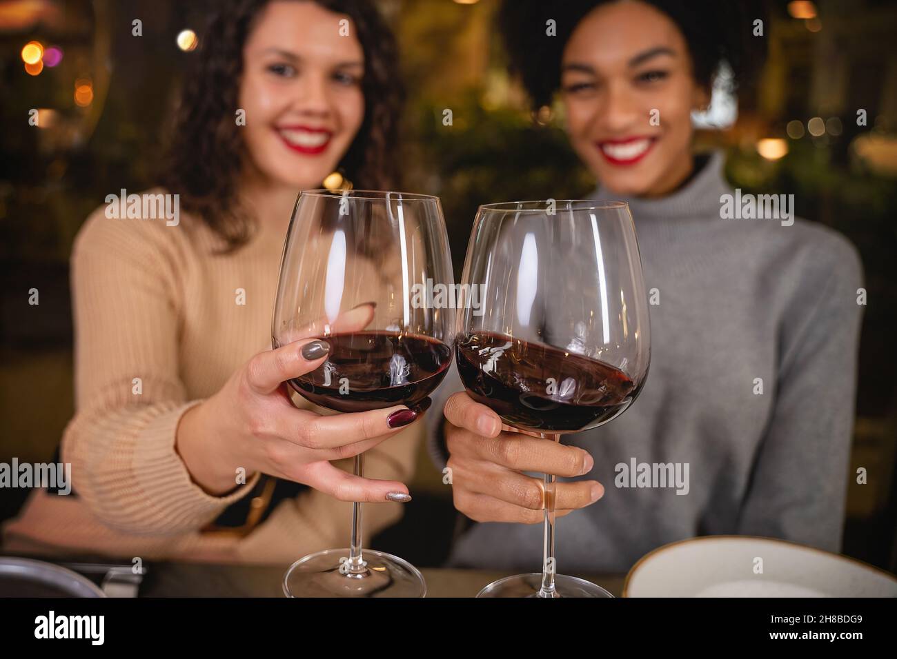 Couple of multicultural girlfriends clebrating toasting red wine at bar ...