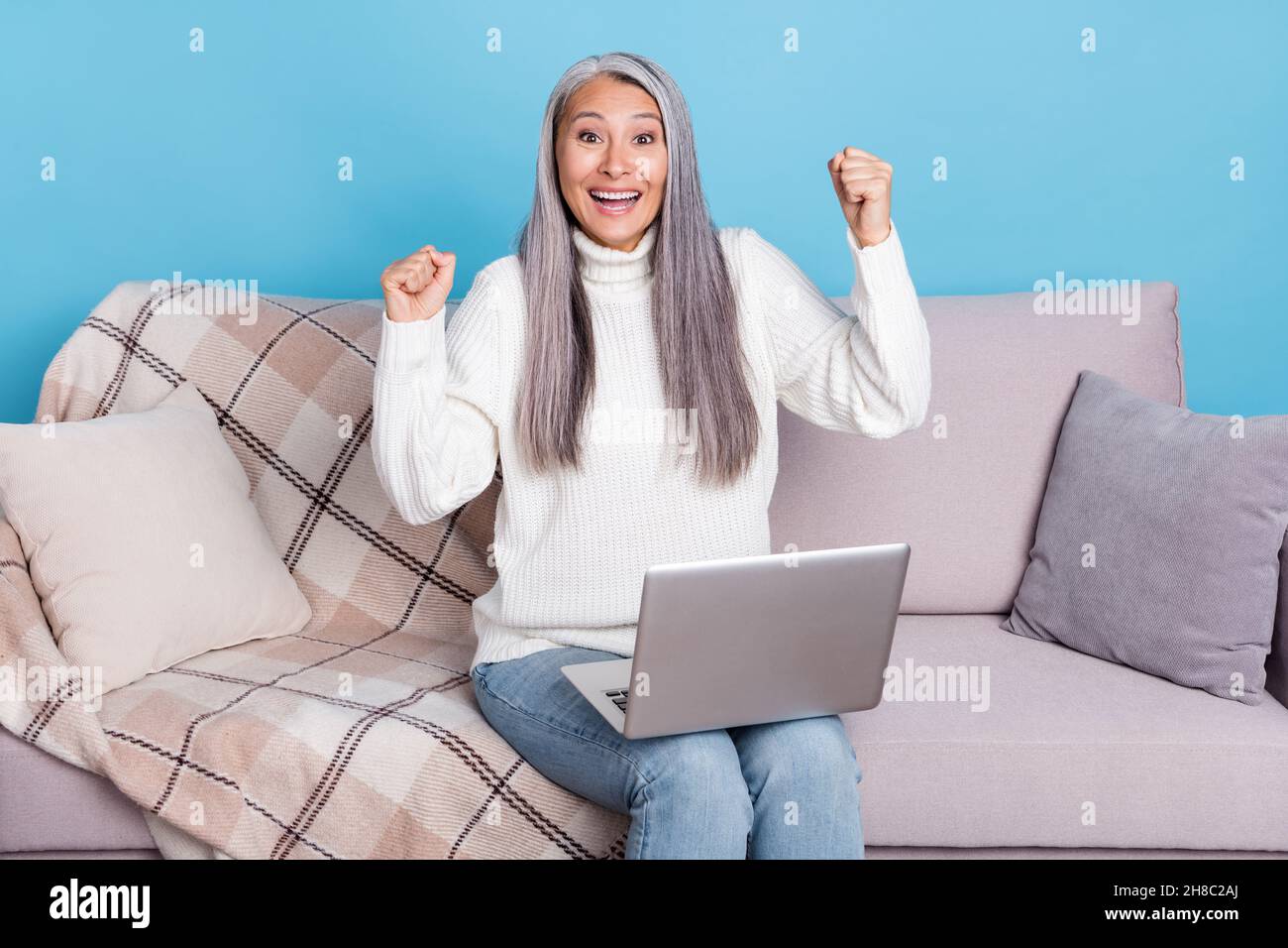 Photo of excited senior woman rejoice victory fists hands sit divan use ...