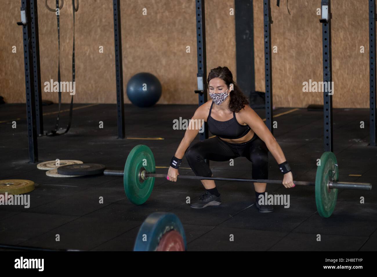 Young sporty woman in a protective mask exercising with a barbell in ...