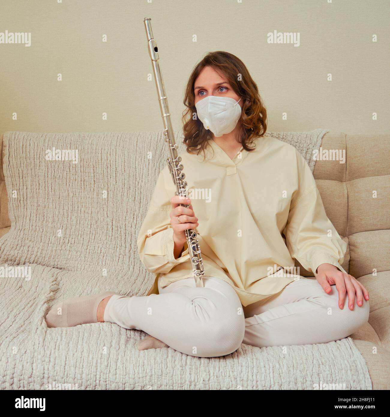 Woman musician with flute in medical face mask playing at home on sofa ...