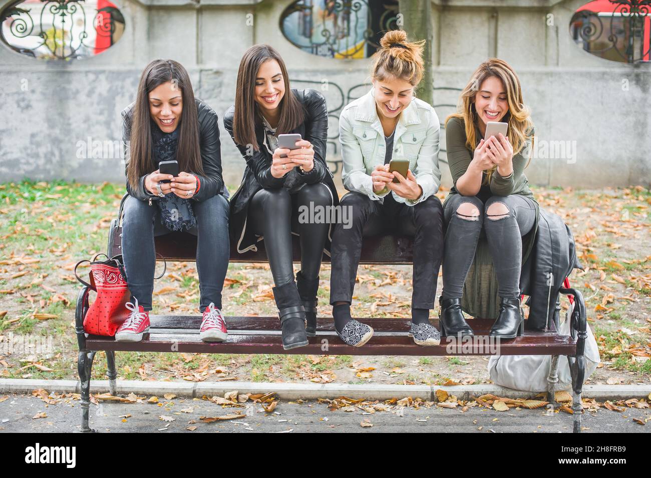 Happy four friends using cellphones on park bench. Every one with his ...