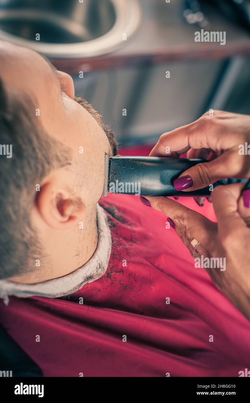 Female barber shaving a client's beard with trimmer in a barber shop ...