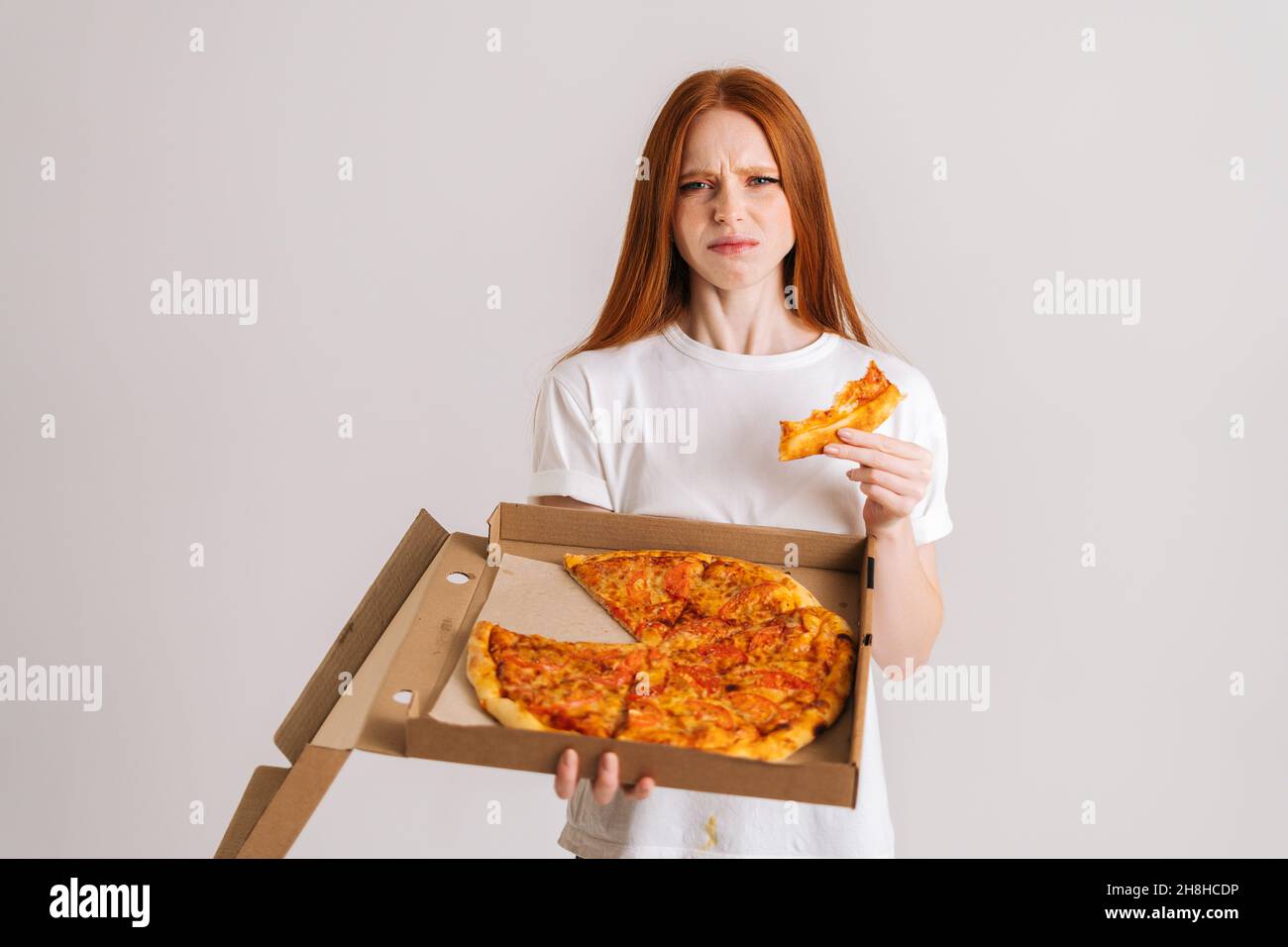 Studio portrait of unhappy young woman eating bad quality pizza holding ...