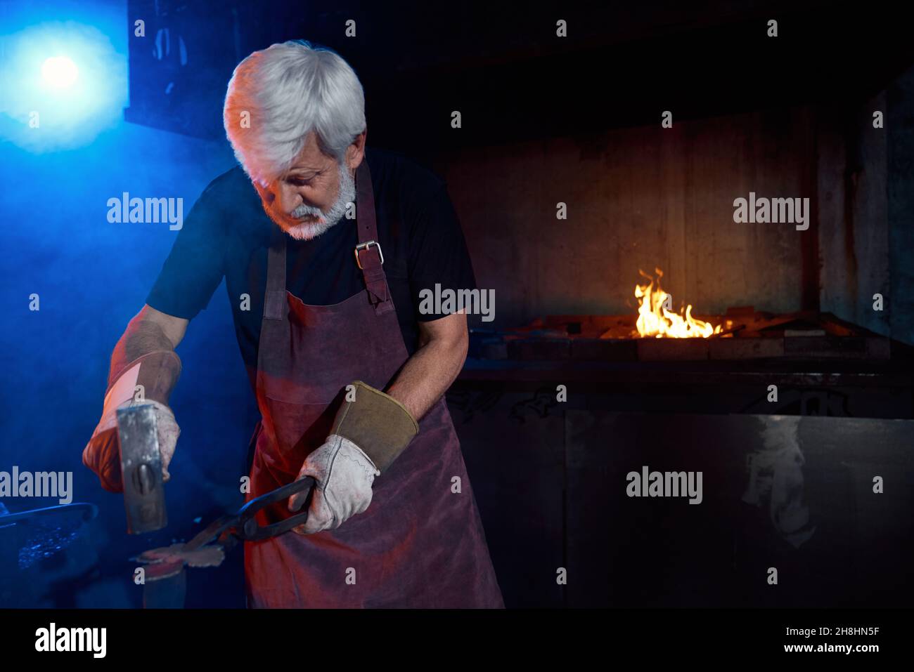 Focused mature man shaping heated steel in safety gloves and apron ...