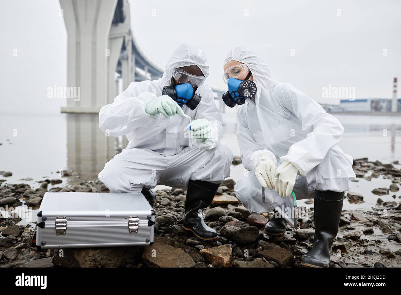 Two people wearing hazmat suits collecting probes by water, toxic waste ...