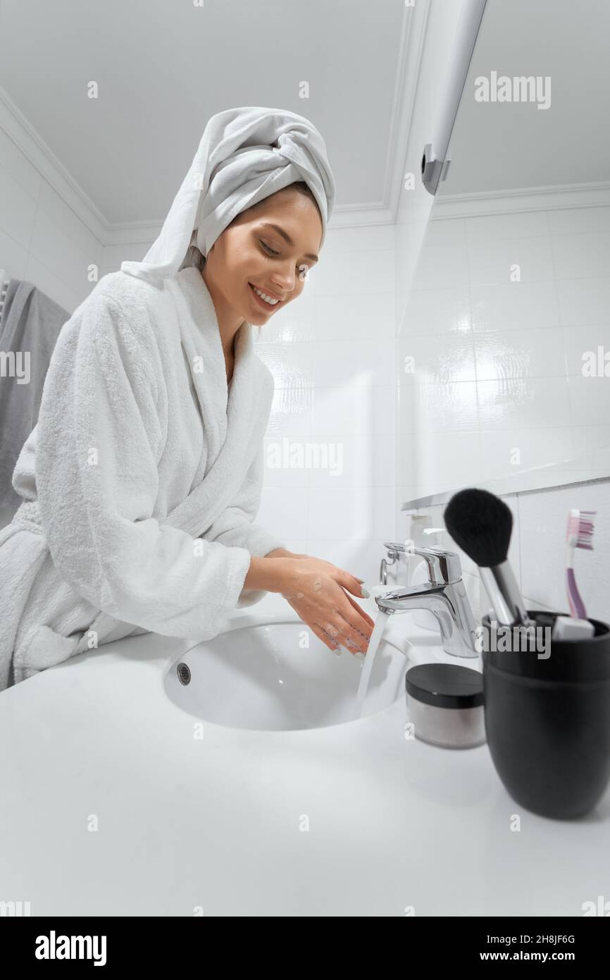 Smiling young woman in white robe washing hands with soap in bathroom ...