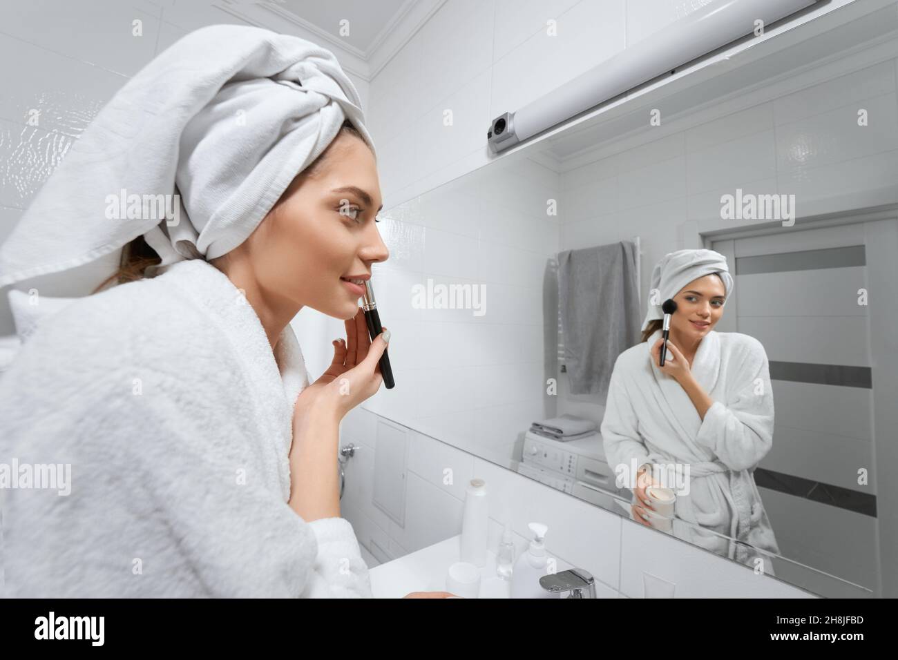 Pretty woman holding blush in hand and doing makeup in hotel bathroom ...