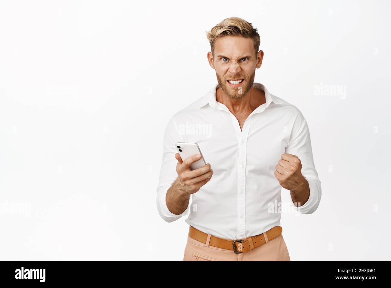 Angry blond man holding cellphone, clenching fist furious and upset ...