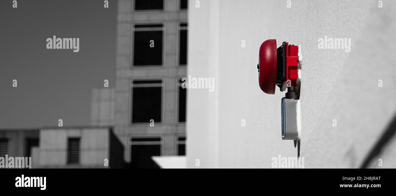 Red alarm bell on white wall with out of focus buildings in background ...