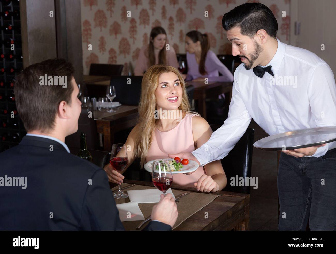 Polite waiter bringing ordered dishes to smiling couple at restaurant ...
