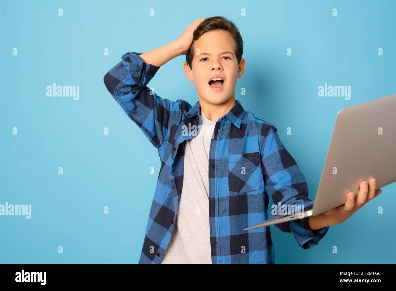 Kid boy shocked holding laptop computer standing over blue background ...