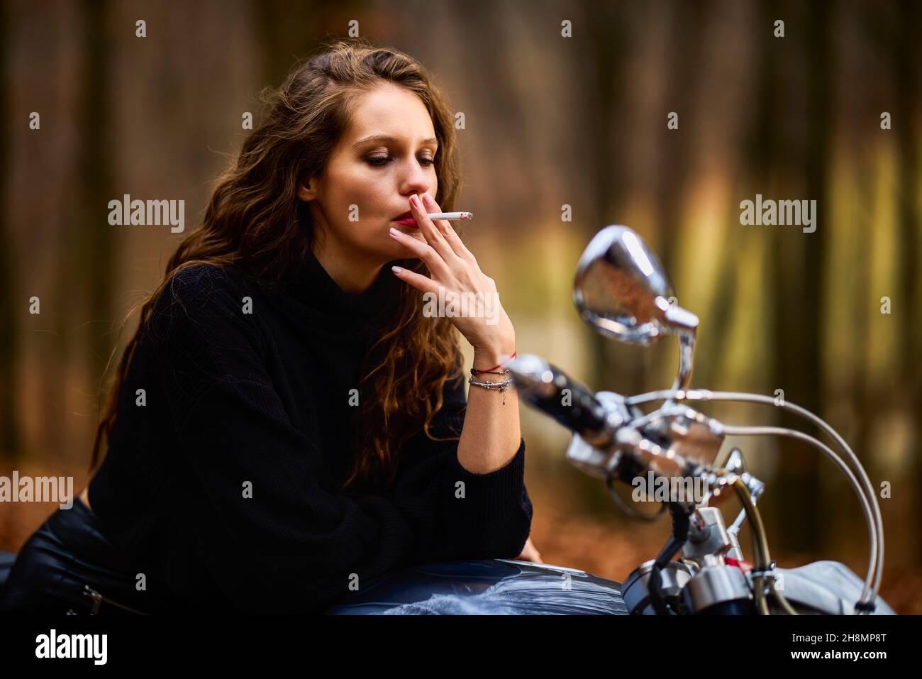 A beautiful long-haired woman smoking on a chopper motorcycle in autumn ...