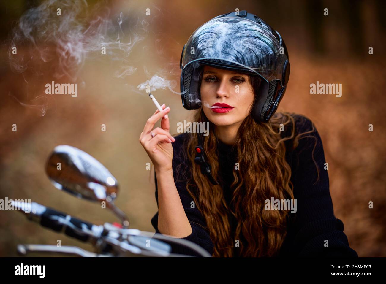 A beautiful long-haired woman smoking on a chopper motorcycle in autumn ...