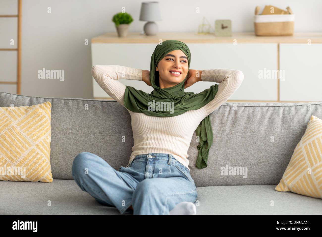 Beautiful young Arab woman in hijab sitting on sofa with hands behind ...