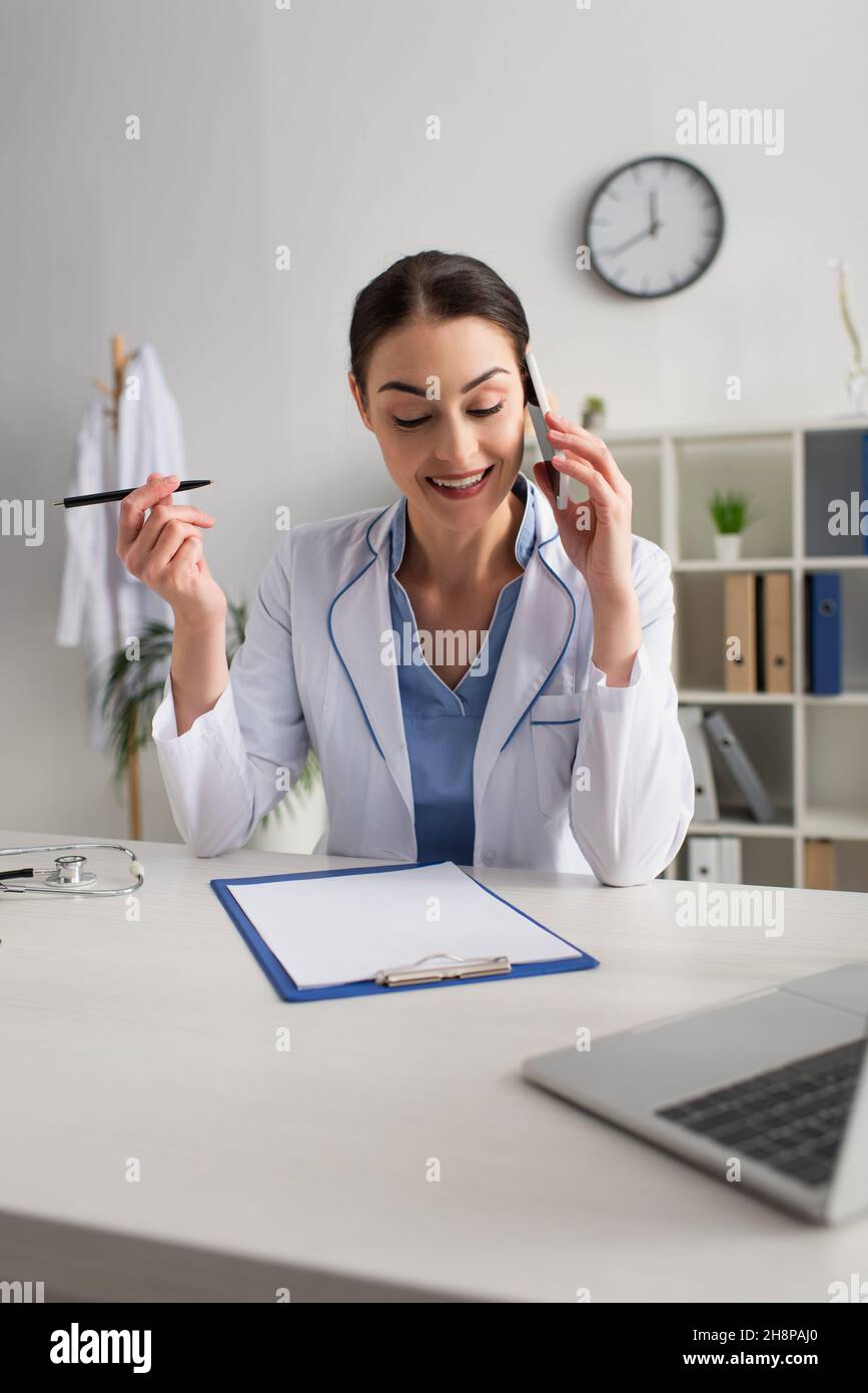 happy doctor holding pen while talking on smartphone near empty ...