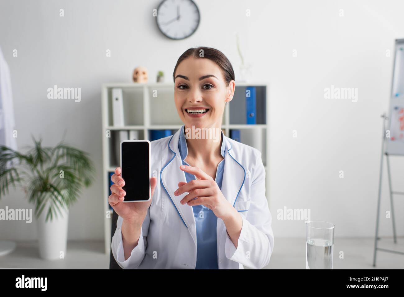 happy brunette doctor pointing at mobile phone with blank screen while ...