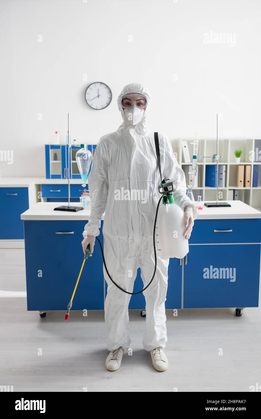 scientist in hazmat suit looking at camera while holding sprayer in ...