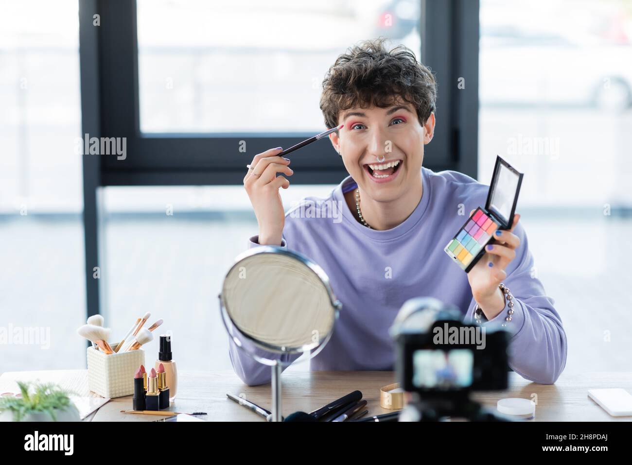 Happy transgender person applying eye shadow and looking a camera near ...