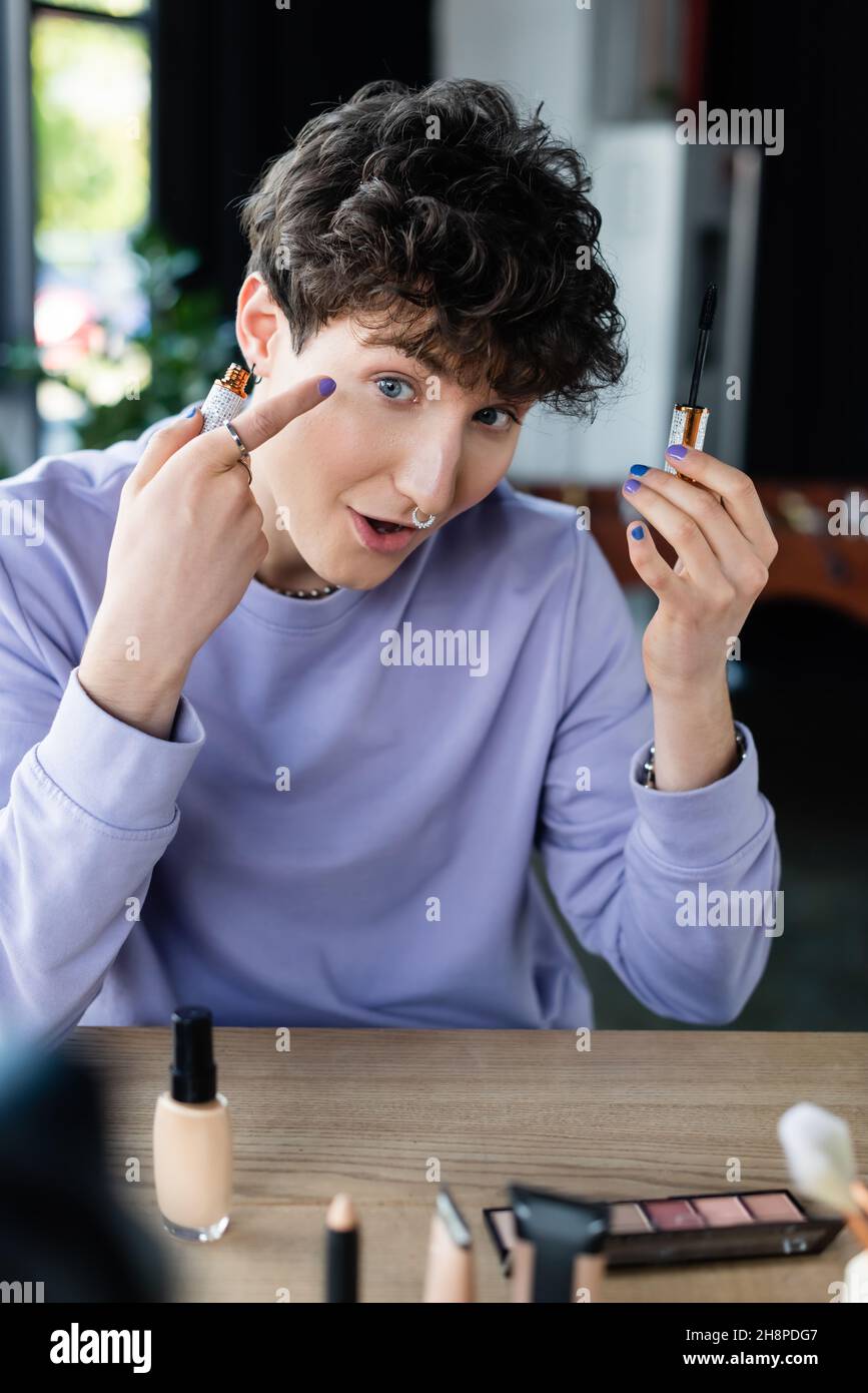 Transgender person holding mascara and pointing at eye near decorative ...