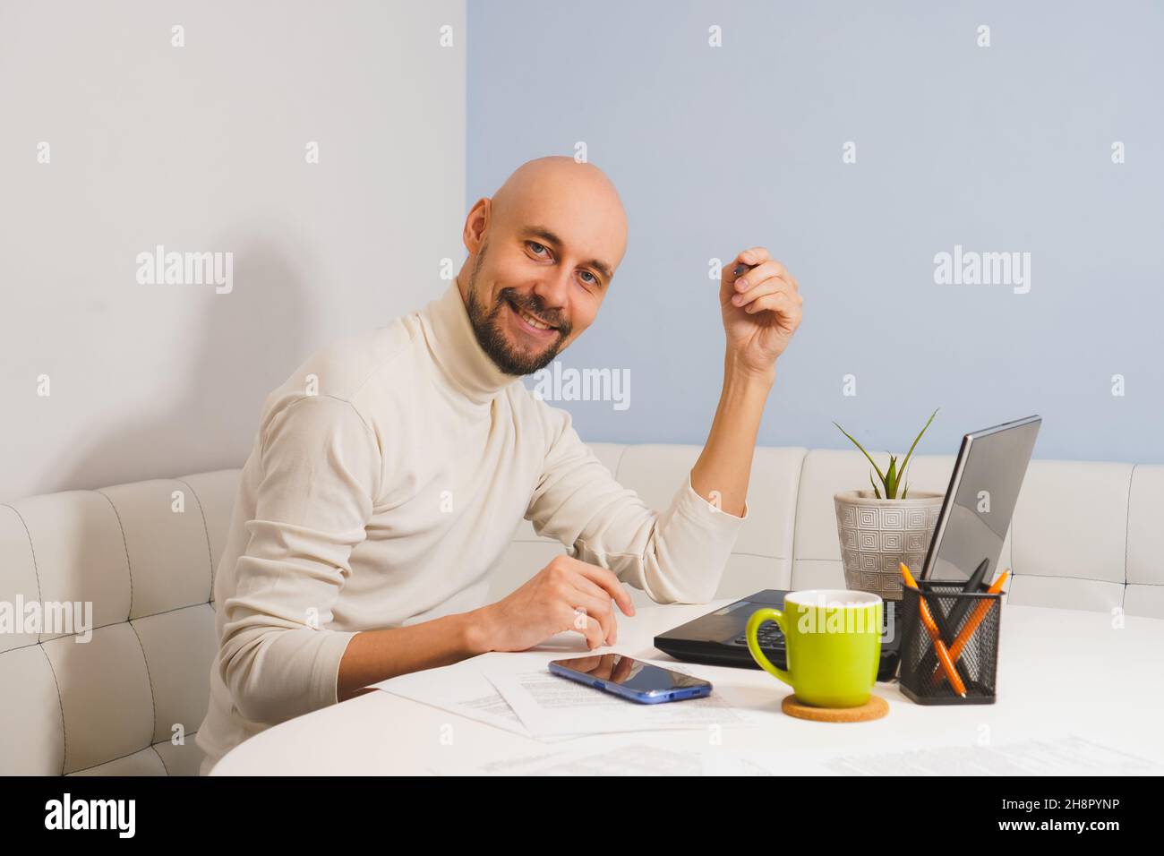Smiling bald man with beard in white turtleneck working on his notebook ...
